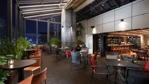 Uma área de refeições ao ar livre com mesas, cadeiras, um bar e vista da cidade à noite.