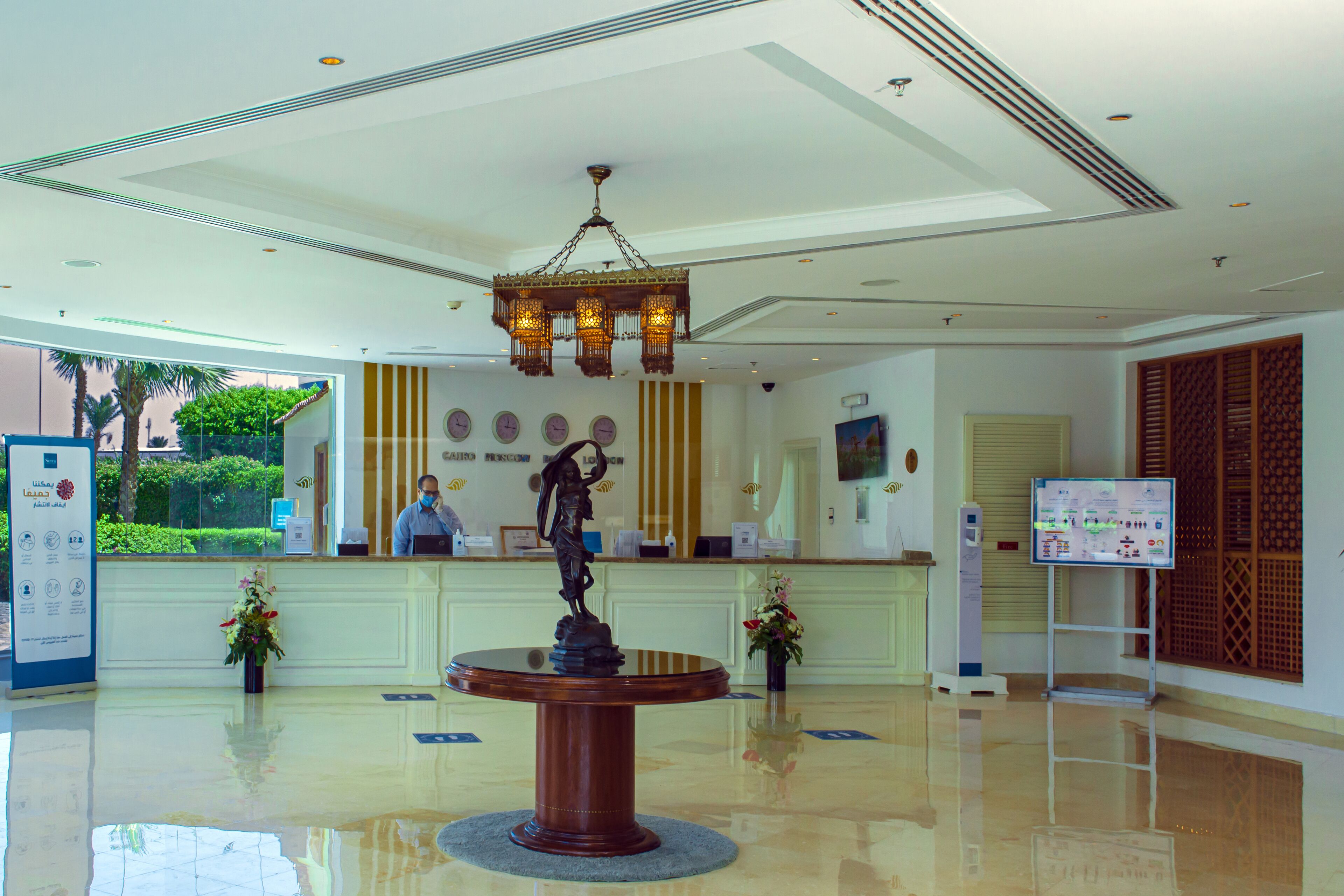 Uma área de recepção com piso lustroso, uma estátua central, um candelabro, um balcão de atendimento e um painel informativo digital.