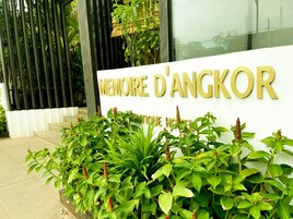 Placa para 'MEMOIRE D'ANGKOR' com fachada de edifício moderno e vegetação exuberante na frente.