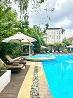 Piscina com espreguiçadeiras e guarda-sóis, rodeada por vegetação e um edifício ao fundo.
