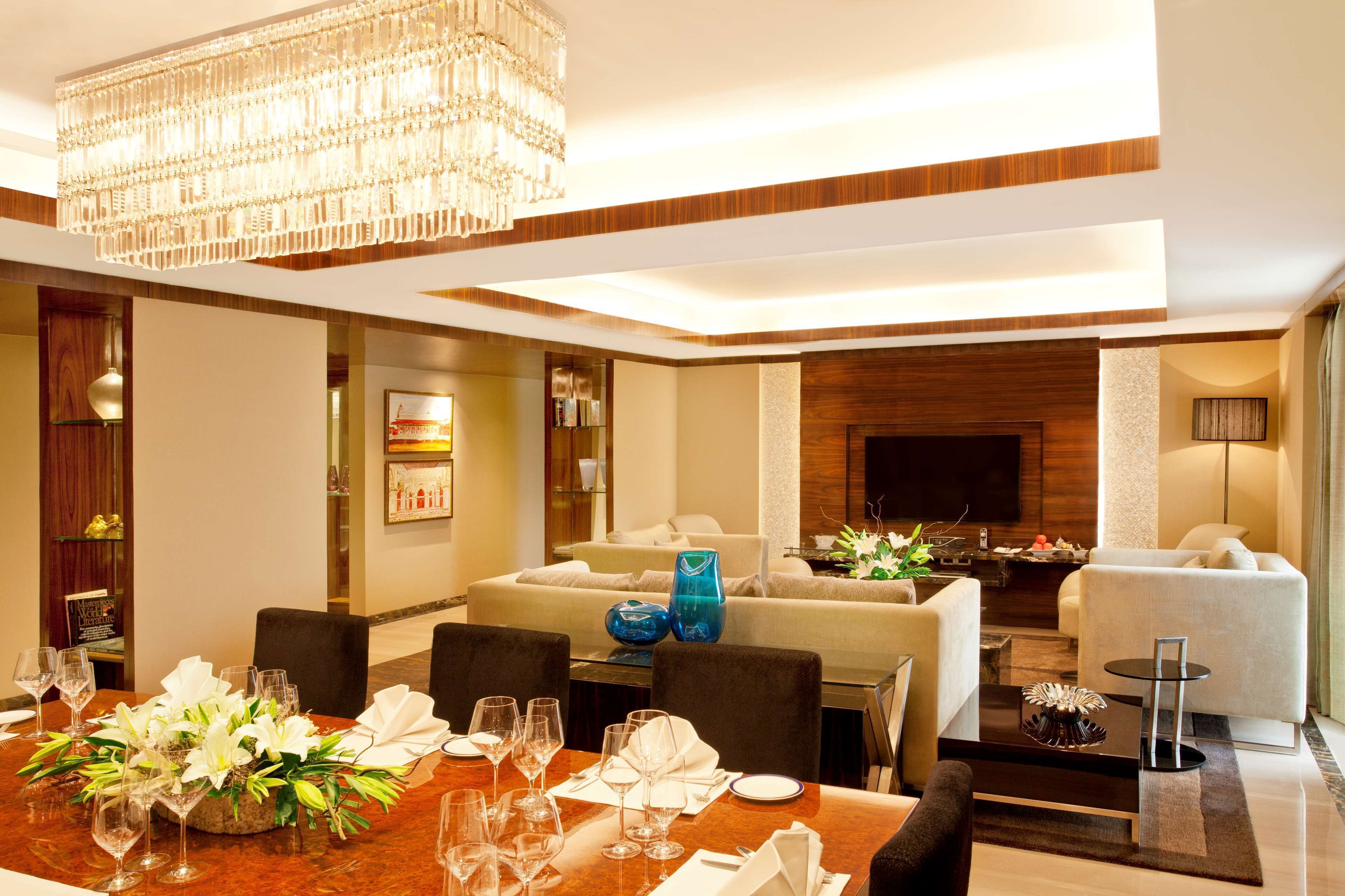 Um espaço de jantar moderno em hotel, com uma mesa de madeira posta para refeição, um candelabro e uma área de estar com sofás e televisão de tela plana.