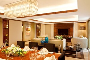 Um espaço de jantar moderno em hotel, com uma mesa de madeira posta para refeição, um candelabro e uma área de estar com sofás e televisão de tela plana.