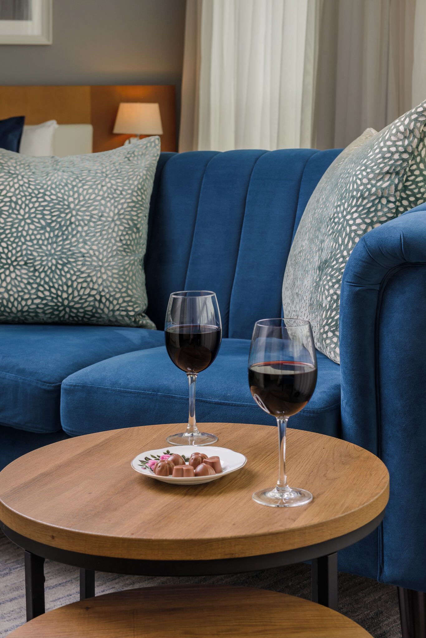 Mesa de madeira redonda com duas taças de vinho e um prato de bombons em um quarto com sofá azul e almofadas estampadas.