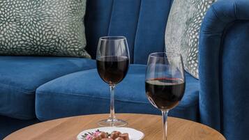 Mesa de madeira redonda com duas taças de vinho e um prato de bombons em um quarto com sofá azul e almofadas estampadas.