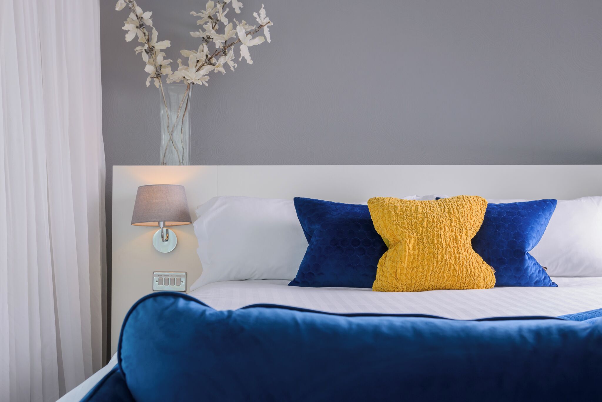 Uma cama bem arrumada com almofadas azuis e amarelas, cabeceira branca e uma luminária na parede.