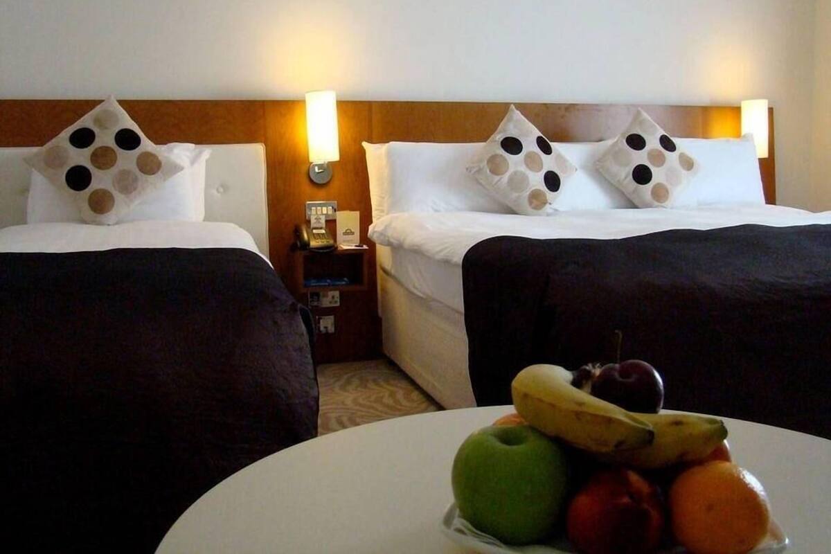 Duas camas com designs de almofadas únicos, uma mesa de cabeceira com abajur e uma tigela de frutas sobre uma mesa.