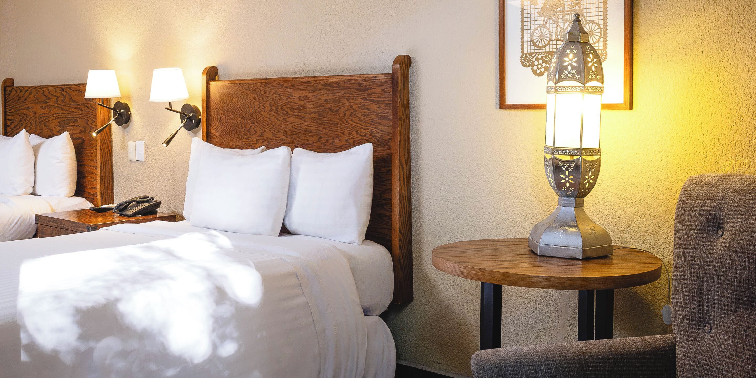 Quarto de hotel com cabeceira de madeira, duas luminárias de apoio, uma mesa de cabeceira redonda de madeira com uma luminária e uma cadeira.