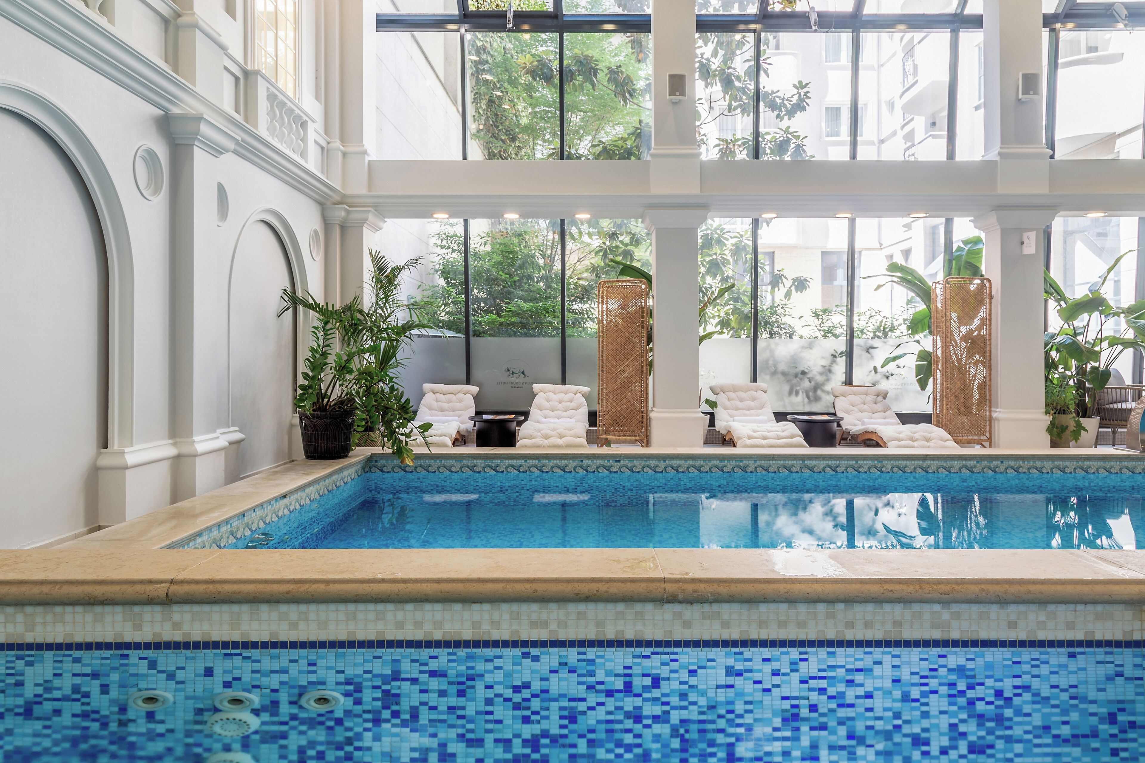 Uma piscina interior com água azul cristalina, rodeada por paredes brancas e grandes janelas.