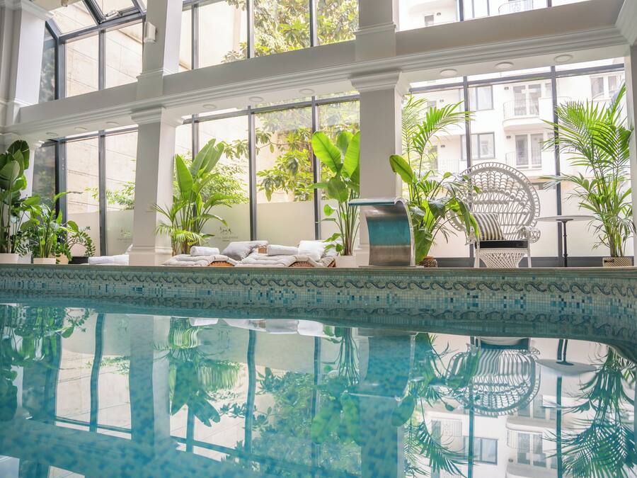 Piscina interna com paredes de vidro transparente, rodeada por plantas em vasos e uma cadeira de vime.
