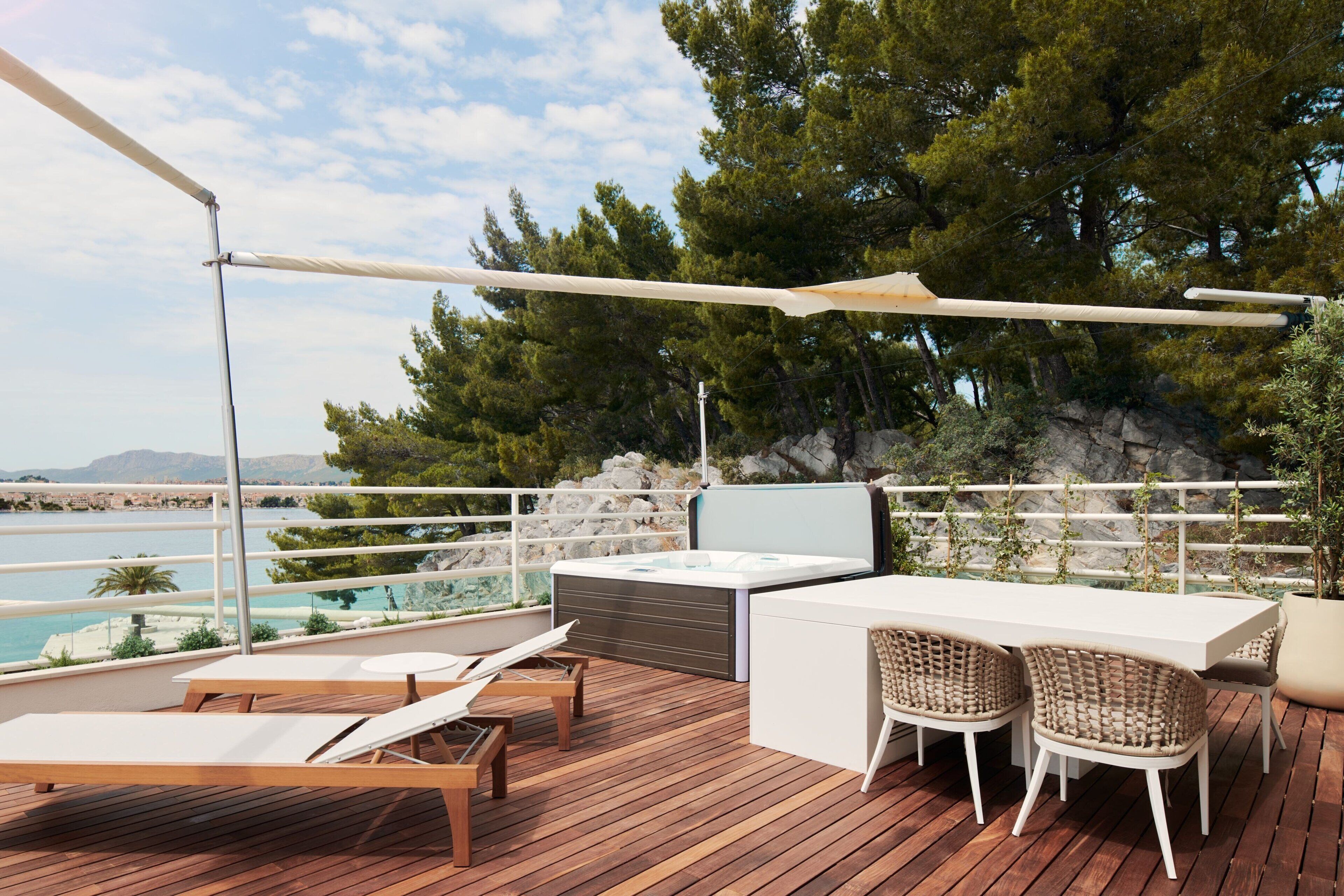 Um deck de madeira com mesa e cadeiras brancas, uma banheira de hidromassagem e vista para o mar e montanhas.