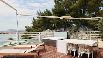 Um deck de madeira com mesa e cadeiras brancas, uma banheira de hidromassagem e vista para o mar e montanhas.