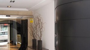 Um lobby moderno com entrada de vidro, uma grande estrutura cilíndrica preta e um arranjo decorativo de plantas.