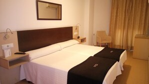 Quarto de hotel com uma cama grande, mesa de cabeceira, cadeira, escrivaninha, espelho e janela com cortinas.