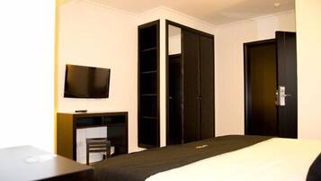 Quarto de hotel com uma cama, uma escrivaninha com cadeira, uma TV na parede e um guarda-roupa.
