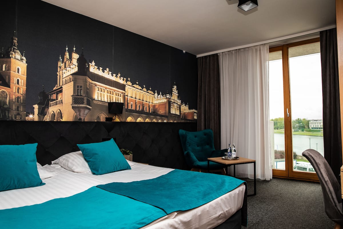 Quarto de hotel com uma cama grande, dois travesseiros azuis, uma pequena mesa com uma garrafa, uma cadeira e vista para um edifício através de uma janela.
