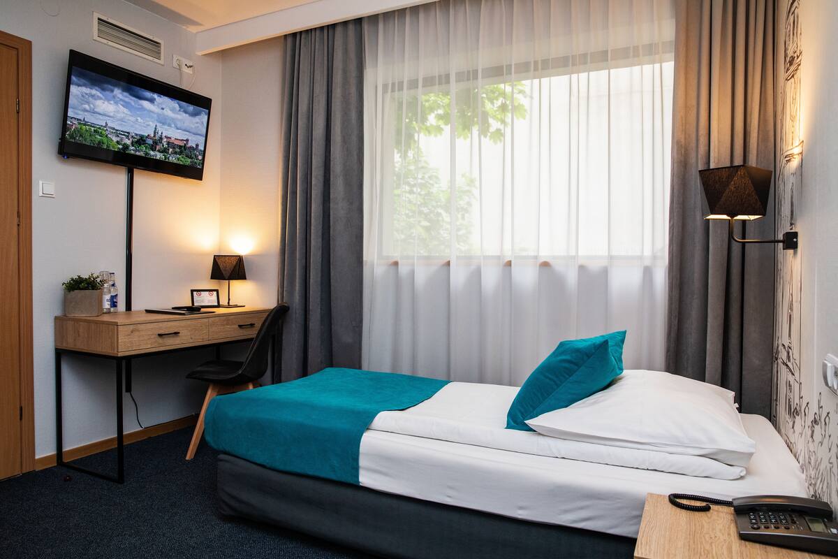 Quarto de hotel com cama, escrivaninha com computador, televisão na parede e janela com cortinas.