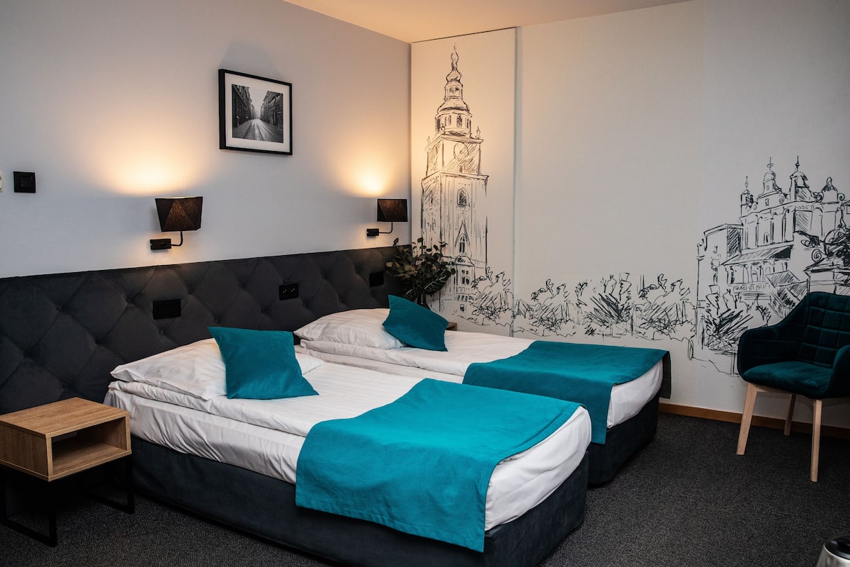 Quarto de hotel com duas camas, cabeceira estofada escura, roupa de cama verde-água, luminária de parede, fotografia emoldurada e um grande mural na parede retratando um edifício.