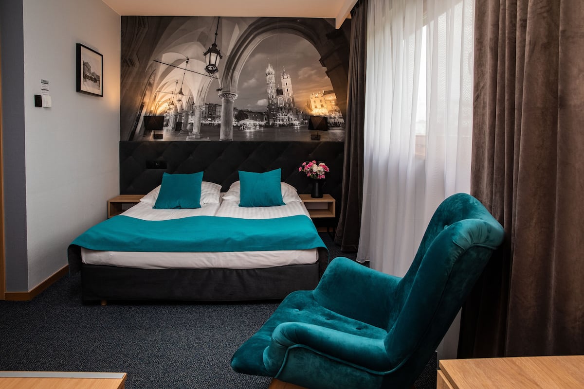 Quarto de hotel com uma cama grande, uma poltrona azul-esverdeada, uma mesa de cabeceira e vista para um edifício histórico pela janela.