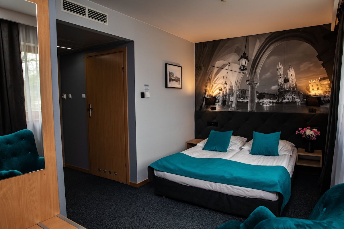 Quarto de hotel com uma cama grande, vista para a cidade através de uma janela em arco, porta de madeira e uma poltrona azul-turquesa.