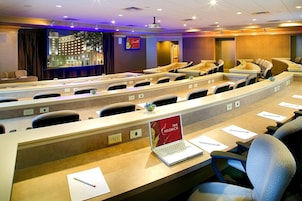 Uma sala de conferências com mesas e cadeiras dispostas em fileiras, uma tela de projetor e um laptop sobre uma das mesas.