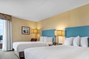 Quarto de hotel com duas camas, uma mesinha de cabeceira, abajur, cadeira e janela com cortinas.