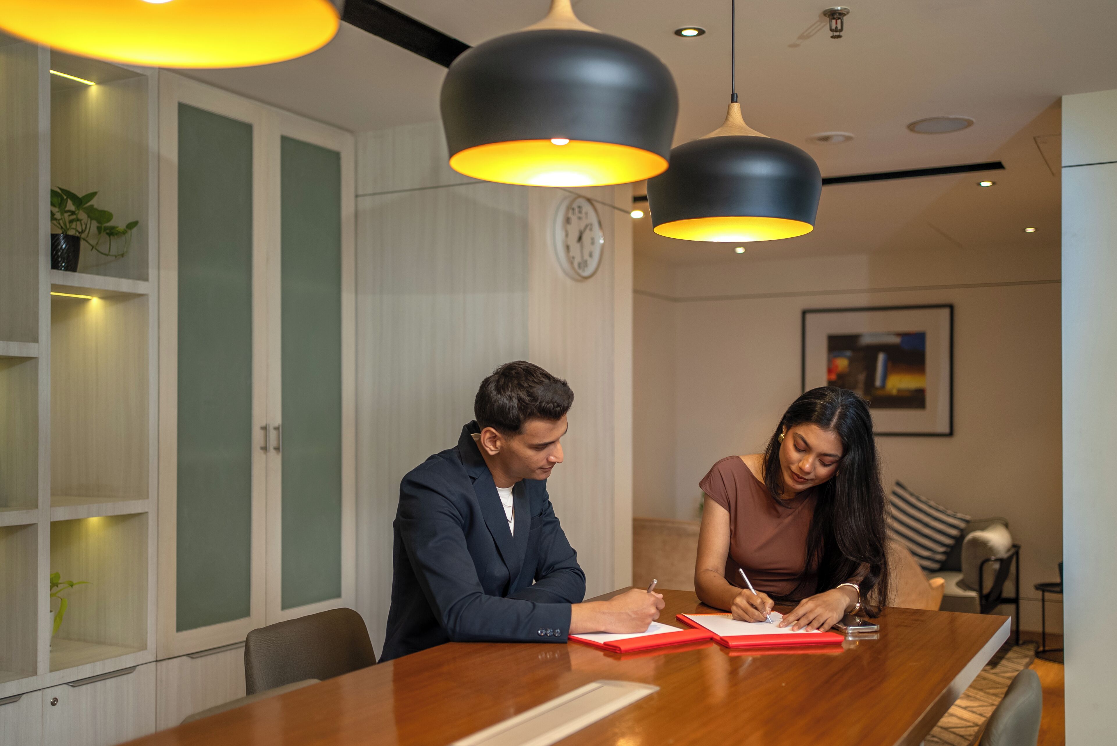 Sala de conferência com mesa de madeira, duas pessoas escrevendo, um relógio e iluminação pendente moderna.