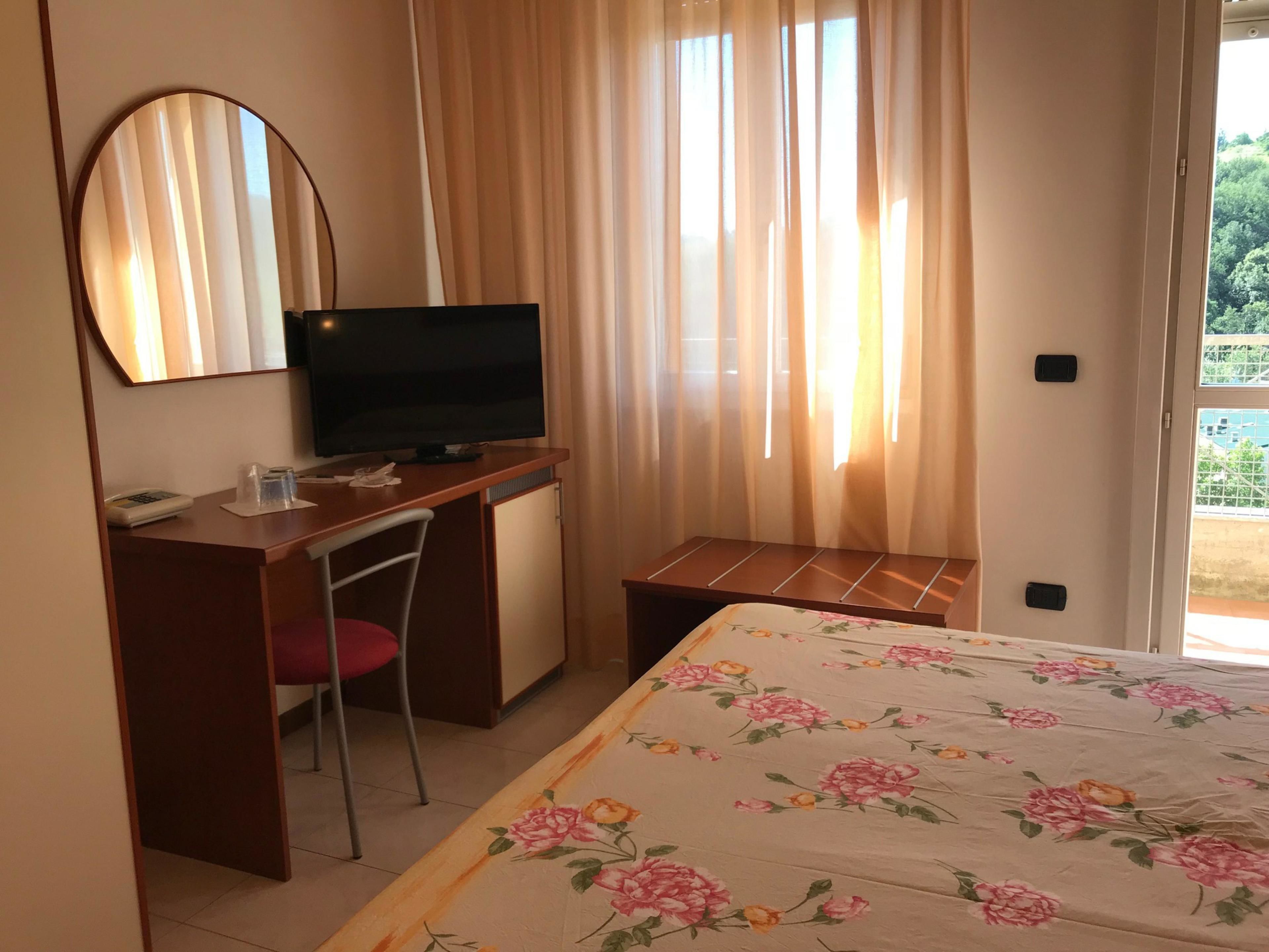 Quarto de hotel com cama, escrivaninha com cadeira, televisão e janela com cortinas.