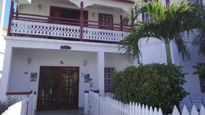 Um edifício de dois andares com fachada branca, uma varanda com guarda-corpo decorativo e uma palmeira em primeiro plano.
