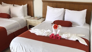 Quarto de hotel com duas camas, cada uma com roupa de cama branca e vermelha, cabeceira de madeira e mesa de cabeceira com um livro e uma luminária.