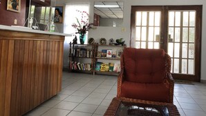 Uma área de recepção com balcão de madeira, uma cadeira vermelha, uma estante com livros e uma janela com vista para o exterior.