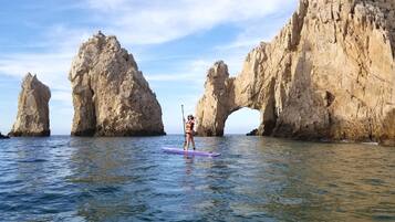 Alguém pratica paddleboarding numa área costeira rochosa, com grandes formações rochosas e um arco natural.