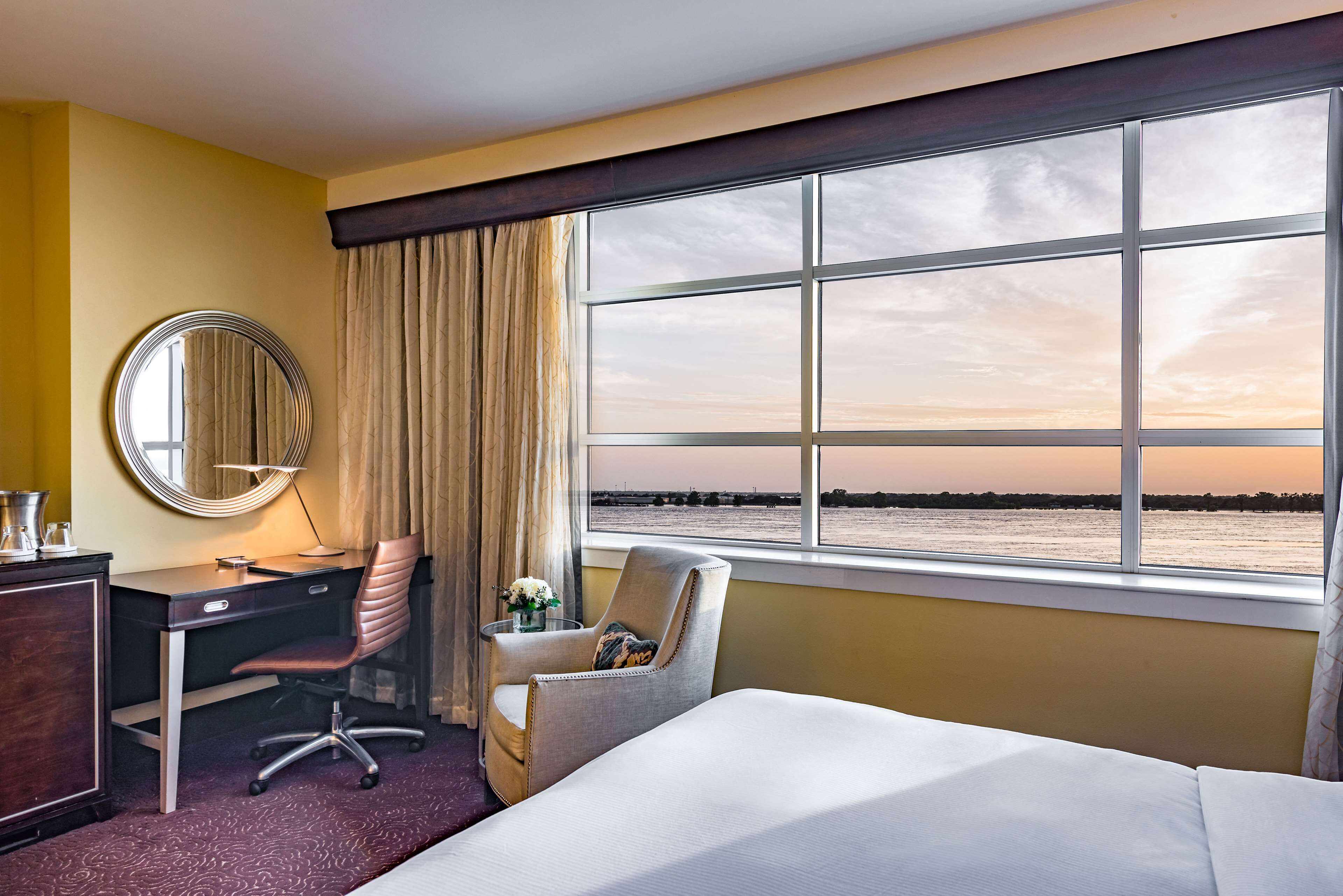 Quarto de hotel com cama, escrivaninha, cadeira e uma janela grande com vista para a água.