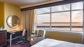 Quarto de hotel com cama, escrivaninha, cadeira e uma janela grande com vista para a água.