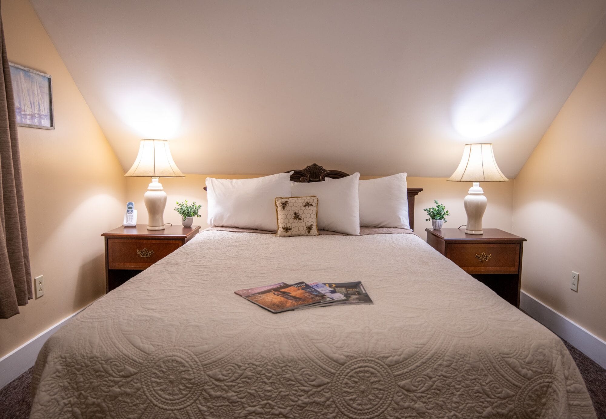 Cama bem arrumada com colcha estampada, duas luminárias de cabeceira, uma mesinha com uma planta e um quadro na parede.
