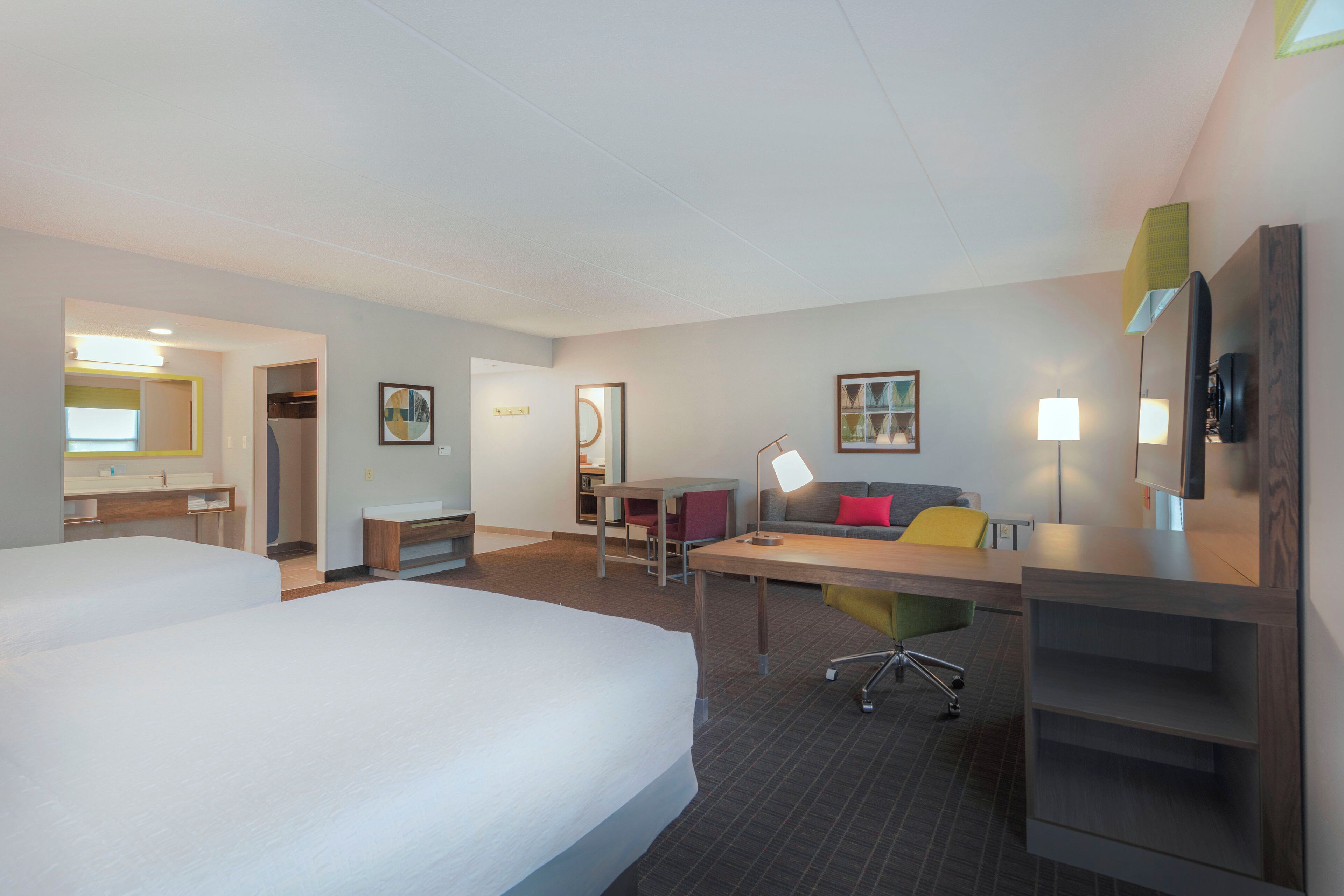 Quarto de hotel com duas camas, uma escrivaninha, uma cadeira, um sofá, uma televisão e um espelho.