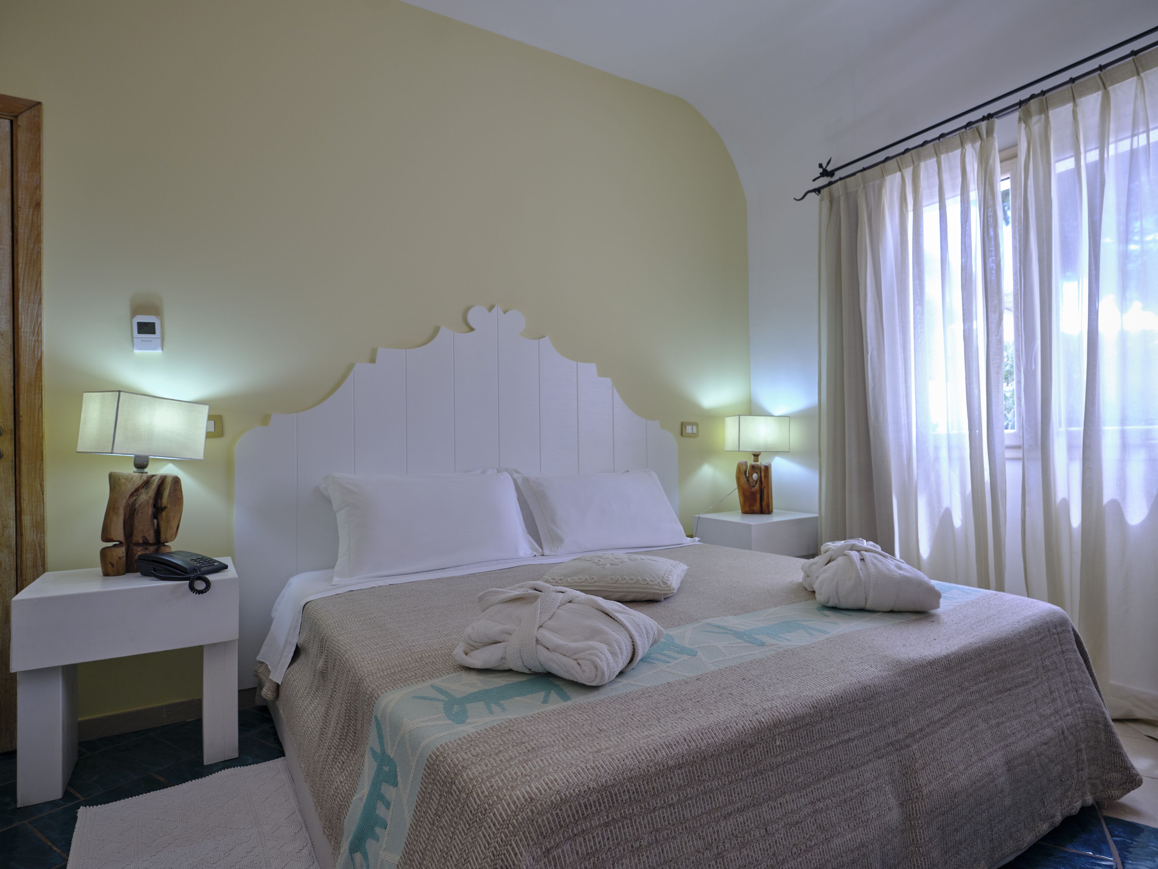 Quarto de hotel bem arrumado, com uma cama, duas mesas de cabeceira, uma luminária e uma janela com cortinas.