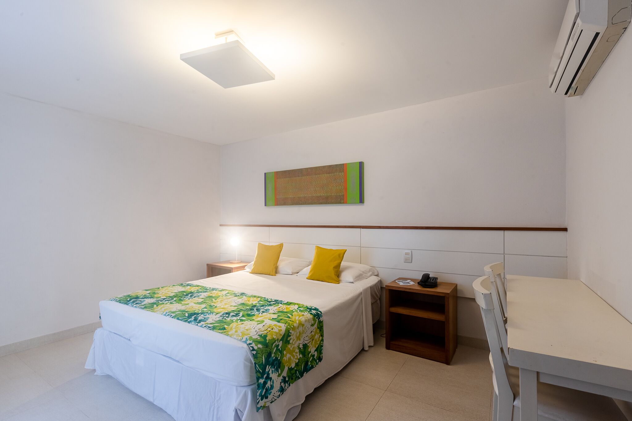 Quarto de hotel com cama, escrivaninha, cadeira, mesa de cabeceira e uma obra de arte na parede.