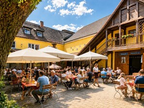Um café ao ar livre com mesas e guarda-sóis, rodeado por edifícios amarelos e uma árvore grande.