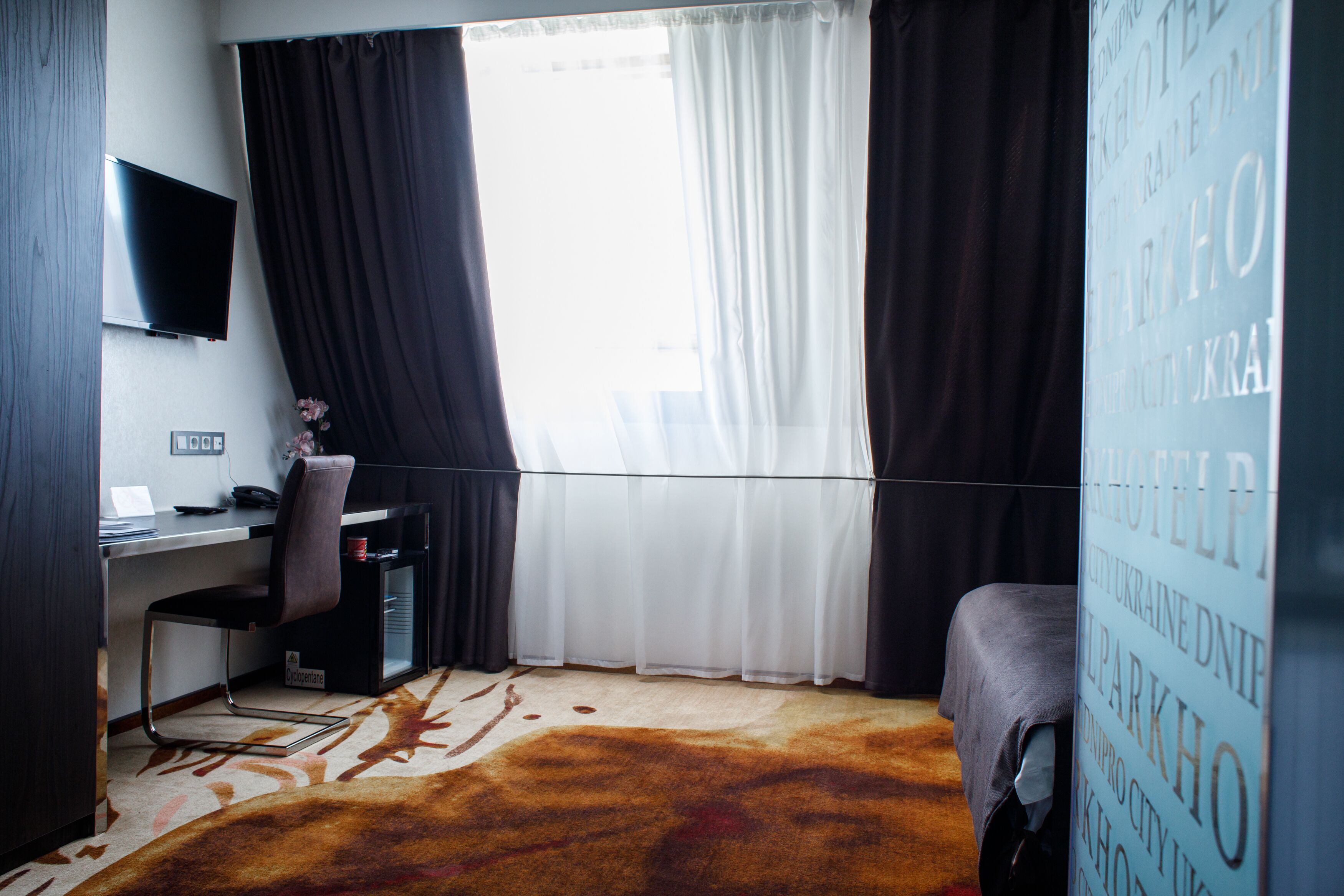 Quarto de hotel com escrivaninha, cadeira e uma janela grande com cortinas leves.