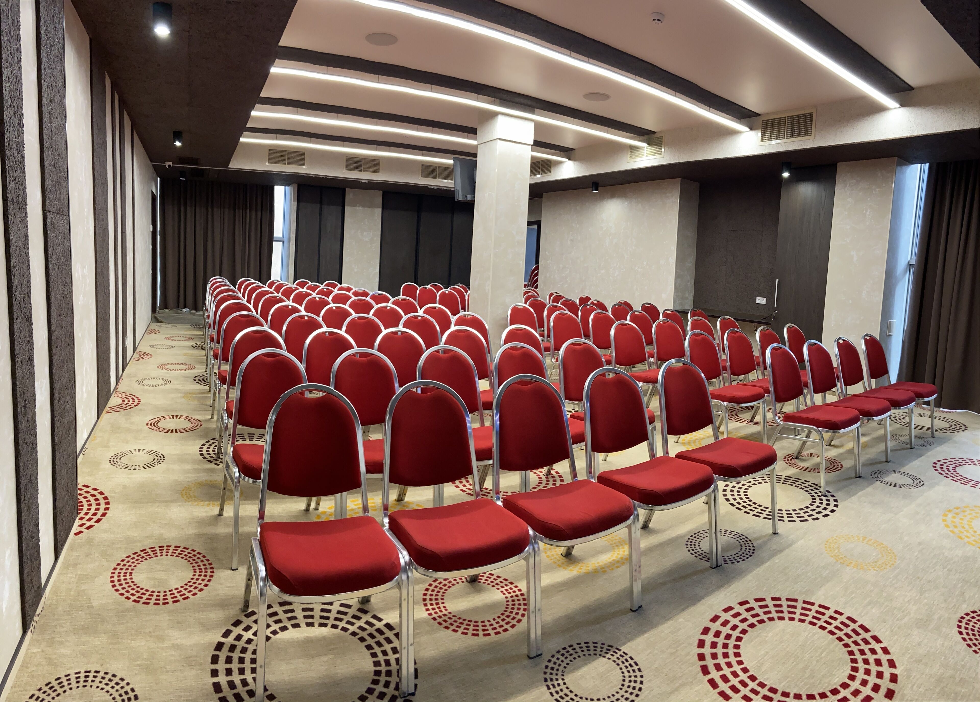 Uma sala de conferências com fileiras de cadeiras vermelhas, carpete estampado e design de teto moderno.