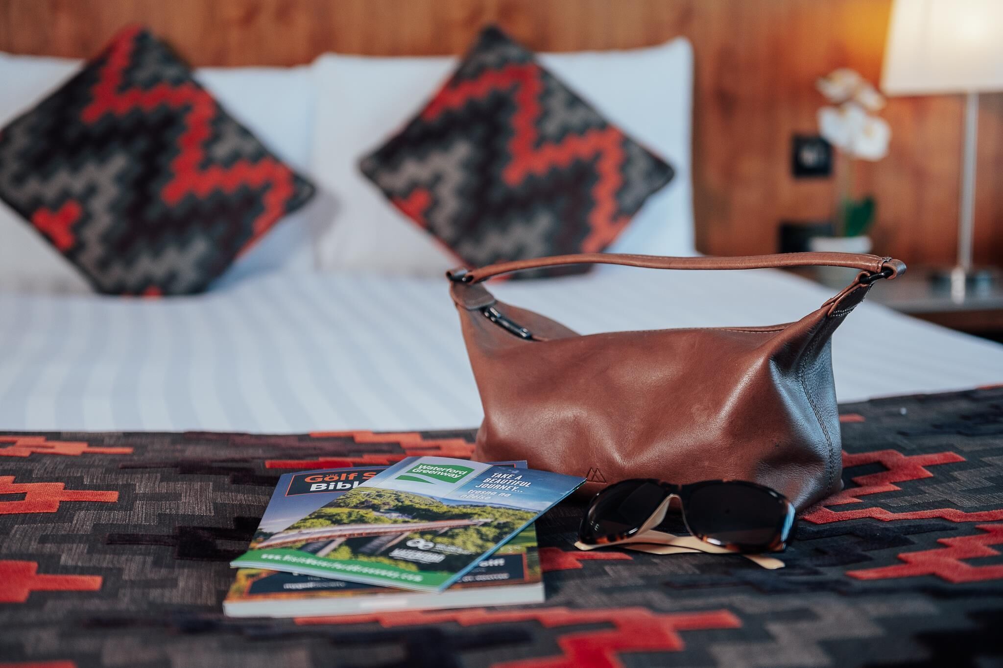 Cama com almofadas estampadas, uma mala de couro marrom, óculos de sol e revistas de viagem sobre um cobreleito.