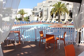 Uma área na piscina com espreguiçadeiras e mesas, cerca branca de piquete e um edifício com varandas ao fundo.