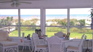 Quarto ensolarado com móveis de vime branco, janelas amplas com vista para a praia e vegetação, e um ventilador de teto.