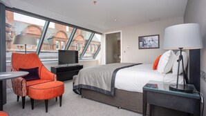 Quarto de hotel moderno com uma cama grande, um espaço de estar com uma poltrona, uma televisão e vista para edifícios por grandes janelas.
