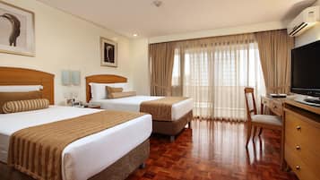 Quarto de hotel com duas camas, uma escrivaninha com televisão, uma cadeira e uma janela grande com cortinas.