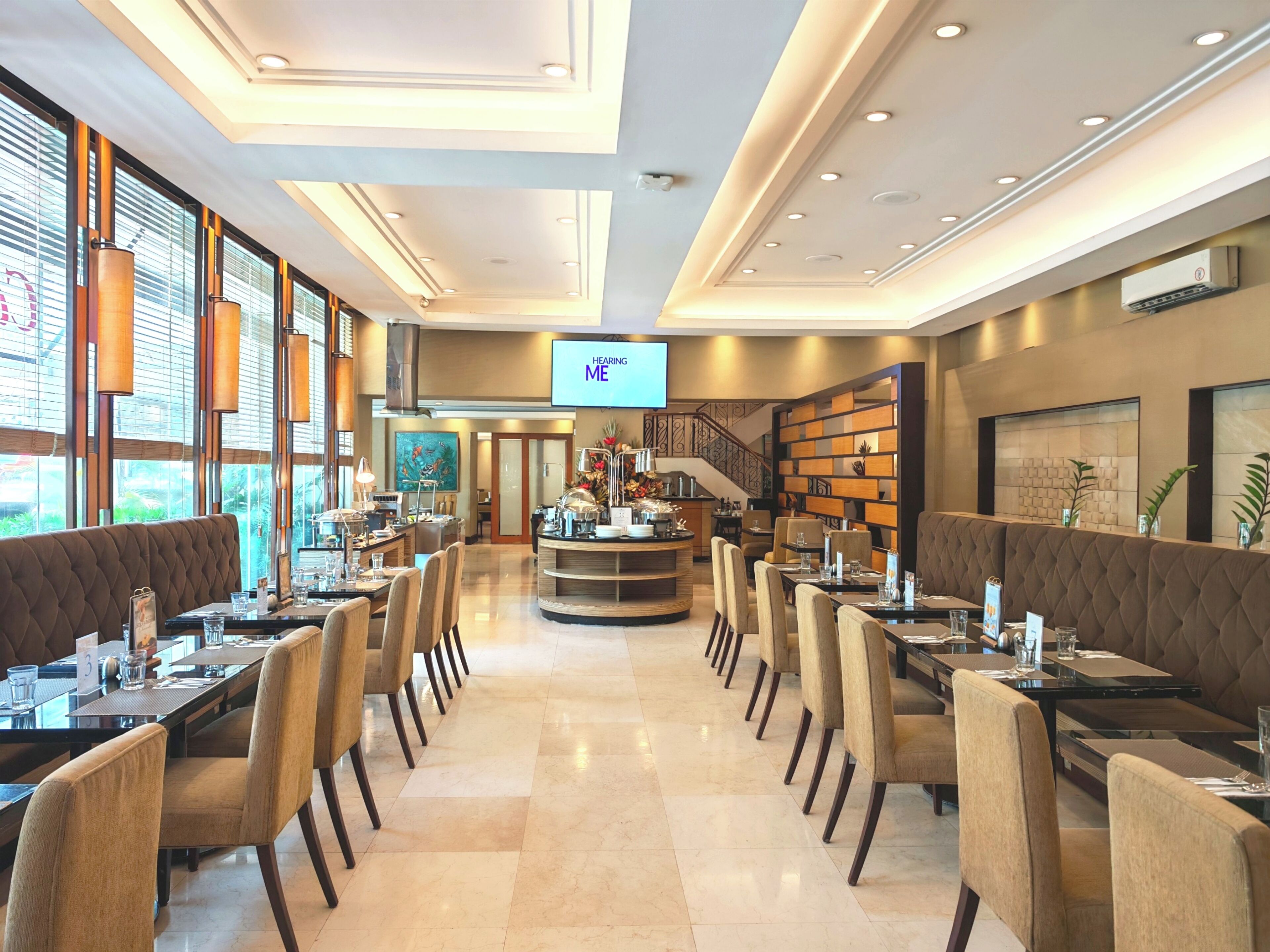 Uma área de refeições de hotel com várias mesas e cabines, um bar central e uma escada que leva a um andar superior.