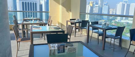 Varanda com mesas e cadeiras, uma mesa com tampo de vidro e vista para um horizonte urbano com edifícios altos.