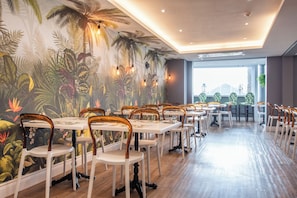 Um restaurante com mesas e cadeiras brancas, um mural tropical e vista para edifícios através de grandes janelas.