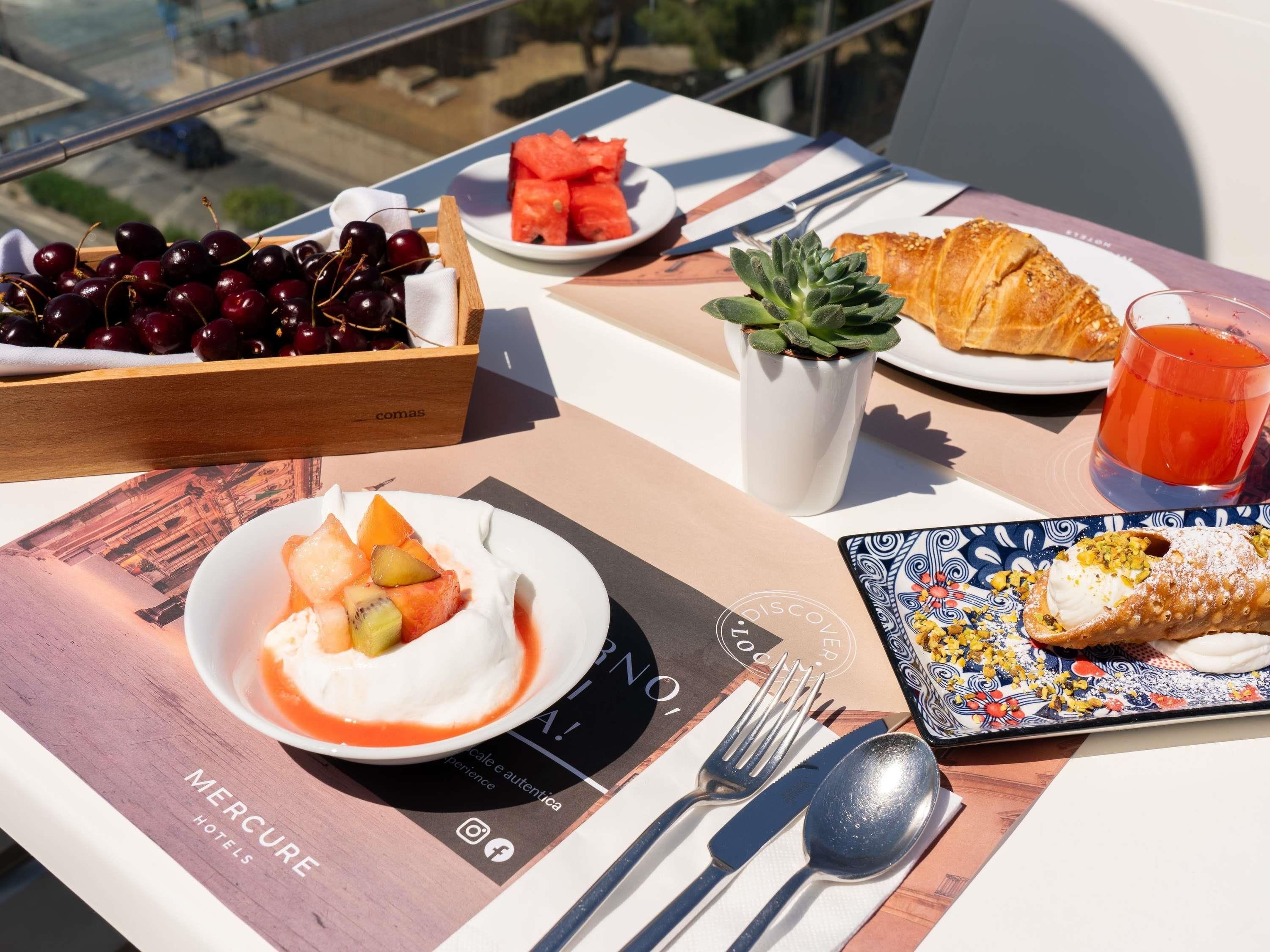 Uma mesa com diversos alimentos, incluindo cerejas, melancia, um croissant e uma sobremesa com frutas e creme.