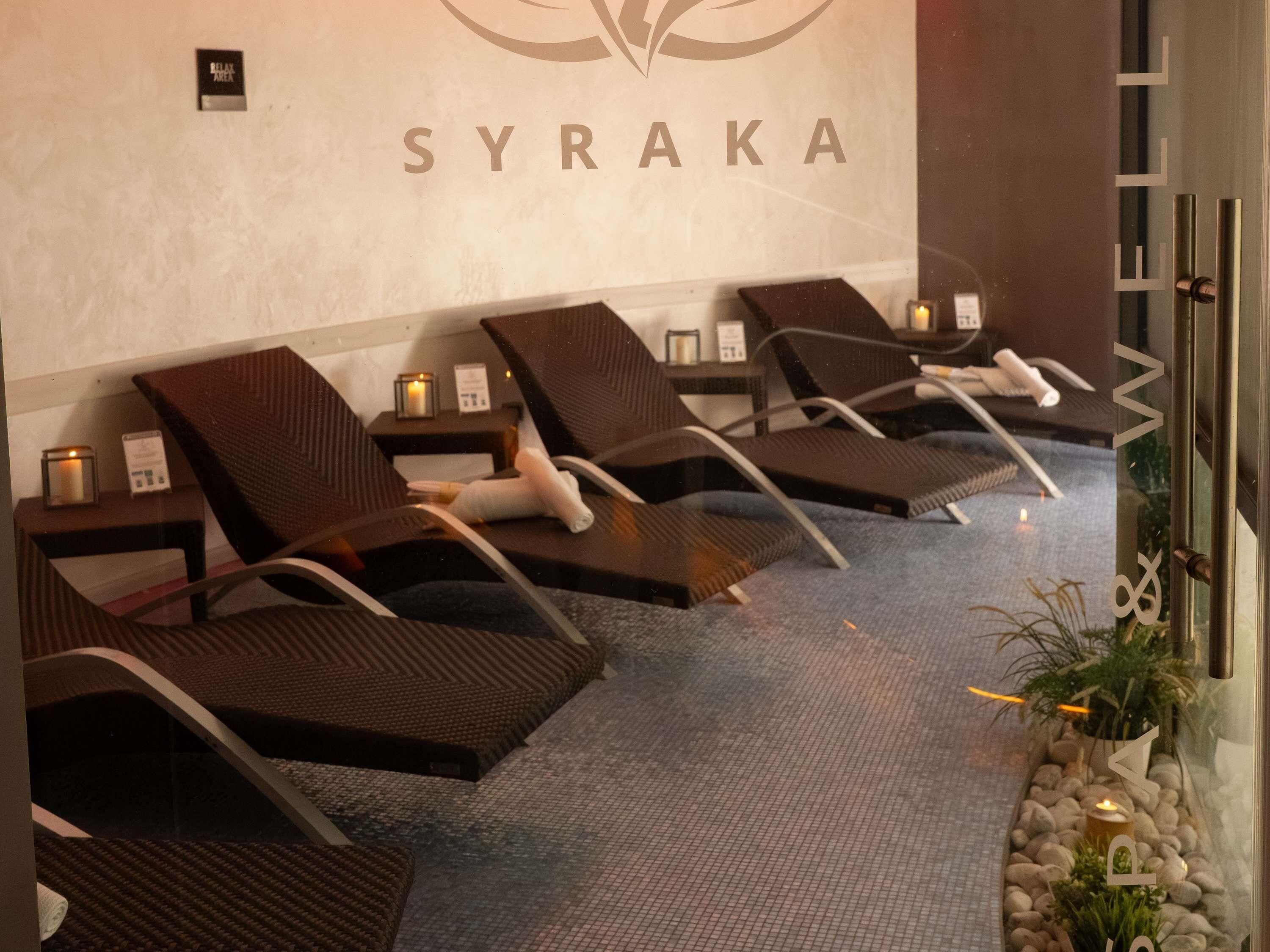 Uma área de spa com cadeiras reclináveis, uma parede com o nome 'SYRAKA' e uma pequena mesa com uma vela.